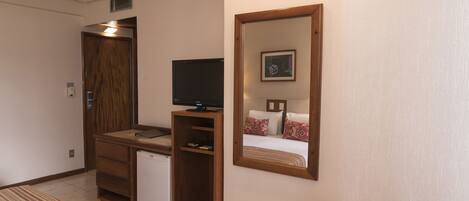 Quarto de hotel compacto com cama, uma pequena mesa com televisão, mini-frigorífico e um espelho na parede.
