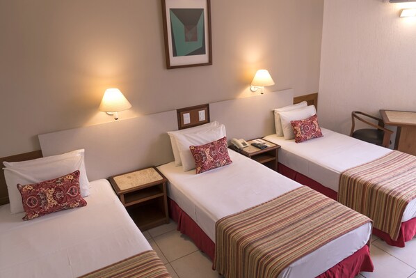 Quarto de hotel com três camas de solteiro, cada uma com almofada estampada em vermelho e branco, mesa de cabeceira de madeira, mesa lateral com luminária e quadro na parede.