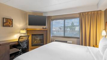 Quarto de hotel com uma cama grande, televisão de tela plana fixada na parede, escrivaninha com cadeira, lareira e janela com cortinas.
