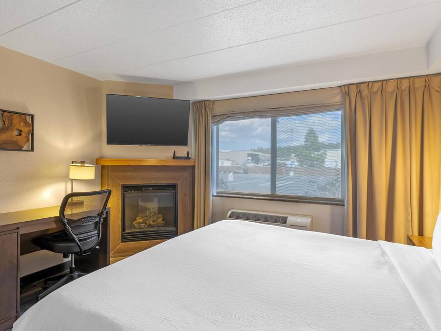 Quarto de hotel com uma cama grande, televisão de tela plana fixada na parede, escrivaninha com cadeira, lareira e janela com cortinas.