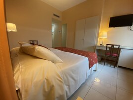Quarto de hotel com cama, escrivaninha, cadeira, televisão e guarda-roupa.