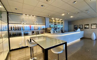 Uma área de bar moderna com balcão de vidro, banquetas e diversas garrafas.