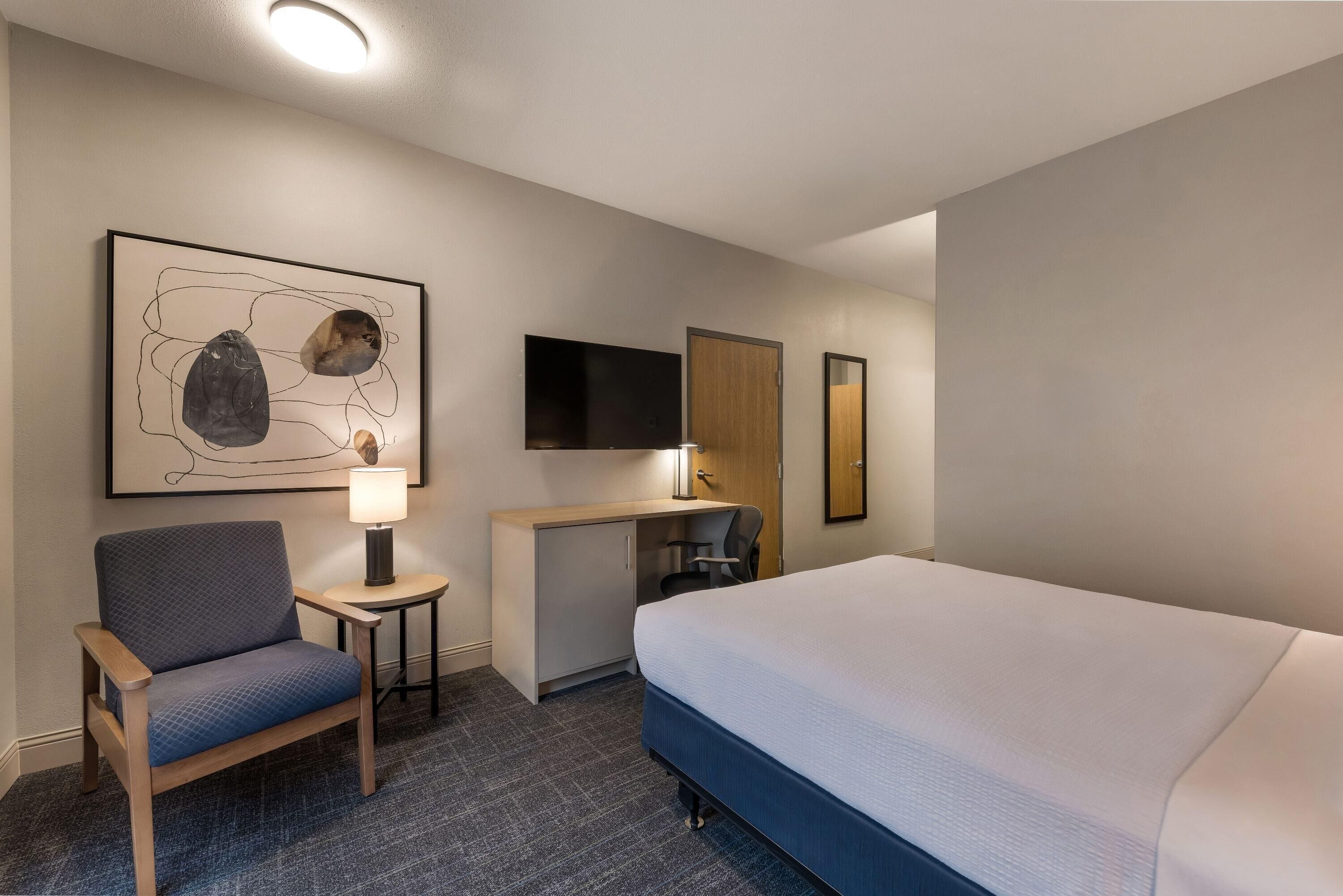 Quarto de hotel com cama, escrivaninha, cadeira, televisão e uma obra de arte emoldurada na parede.