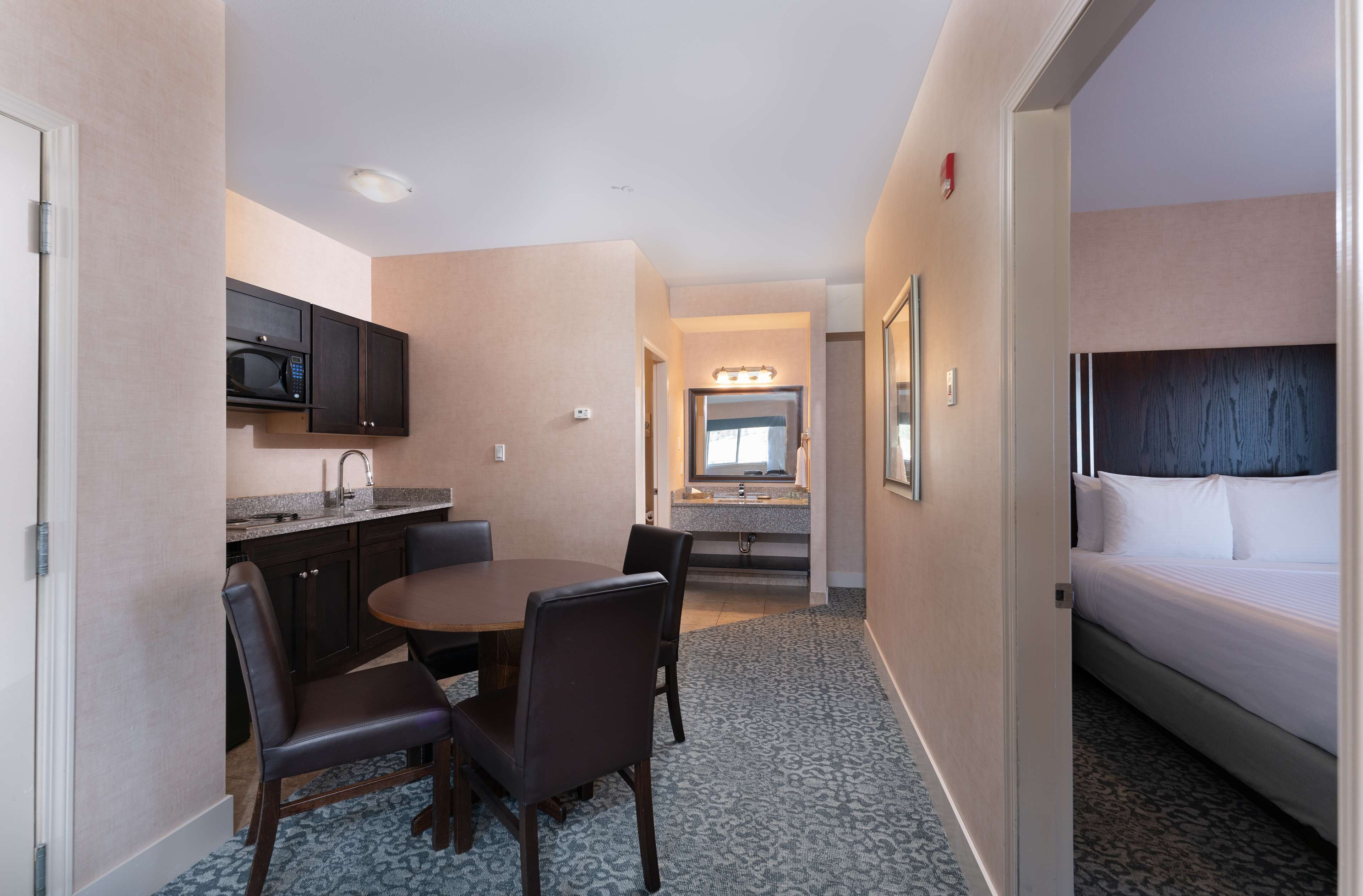 Quarto de hotel com cama, banheiro com pia e espelho, pequena cozinha com micro-ondas e área de jantar com mesa e cadeiras.