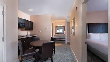 Quarto de hotel com cama, banheiro com pia e espelho, pequena cozinha com micro-ondas e área de jantar com mesa e cadeiras.