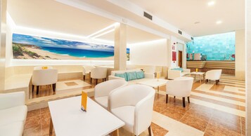 Um saguão moderno de hotel com um mural vista para a praia, móveis brancos e um balcão de recepção.