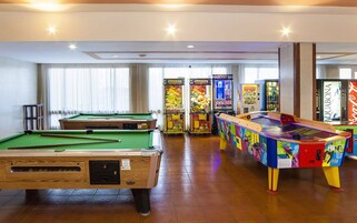Uma mesa de bilhar, máquinas de fliperama e uma mesa de tênis de mesa colorida em um ambiente com piso de madeira e janelas grandes.