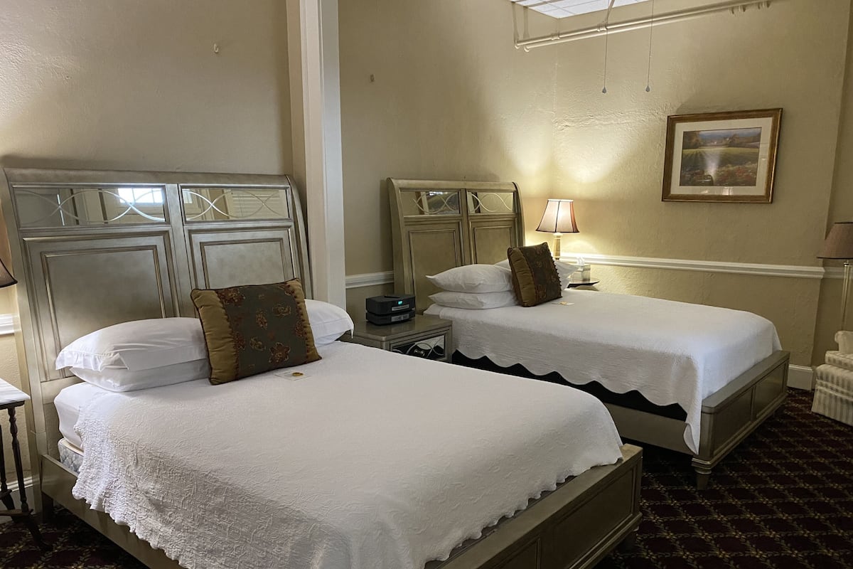 Quarto de hotel com duas camas, cada uma com cabeceira, roupa de cama branca e uma pequena mesa de cabeceira com abajur.