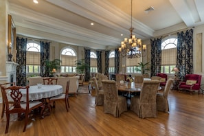 Uma ampla sala de jantar com mesa e cadeiras de madeira, cercada por janelas grandes e um candelabro.