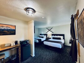 Quarto de hotel com duas camas, uma mesa com micro-ondas, uma lixeira e uma pintura de uma montanha na parede.