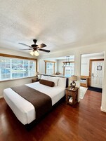 Quarto de hotel com uma cama grande, um ventilador de teto, uma janela com persianas, piso de madeira e uma porta com espelho.