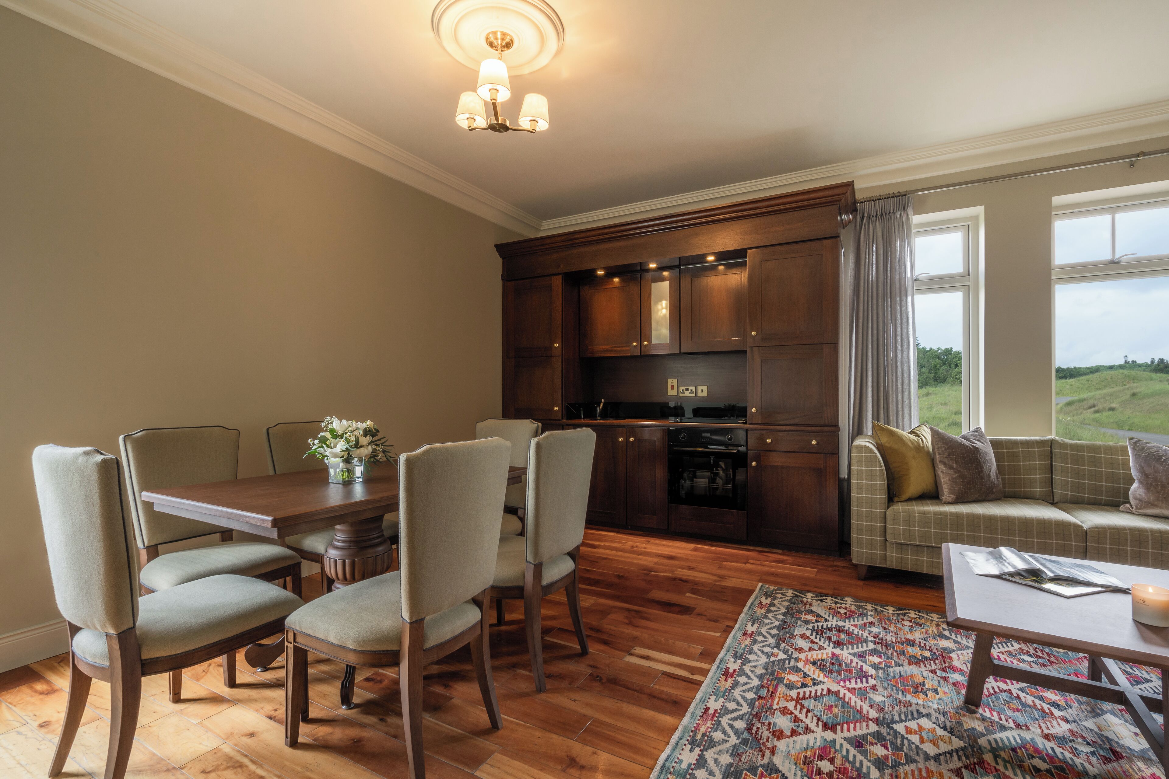 Uma sala de jantar com mesa e cadeiras de madeira, uma cozinha com armários de madeira escura e vista para uma paisagem verde pela janela.