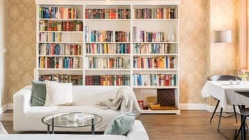 Uma sala de estar com um sofá branco, uma mesa de centro de vidro, uma estante repleta de livros e um espaço de jantar com mesa e cadeiras.