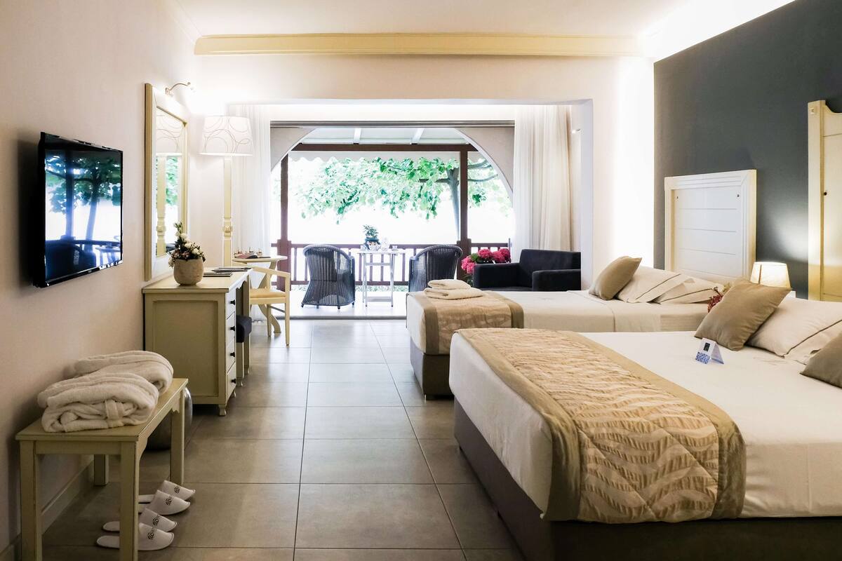 Quarto de hotel com duas camas, televisão, escrivaninha com cadeira e varanda com vista para árvores.