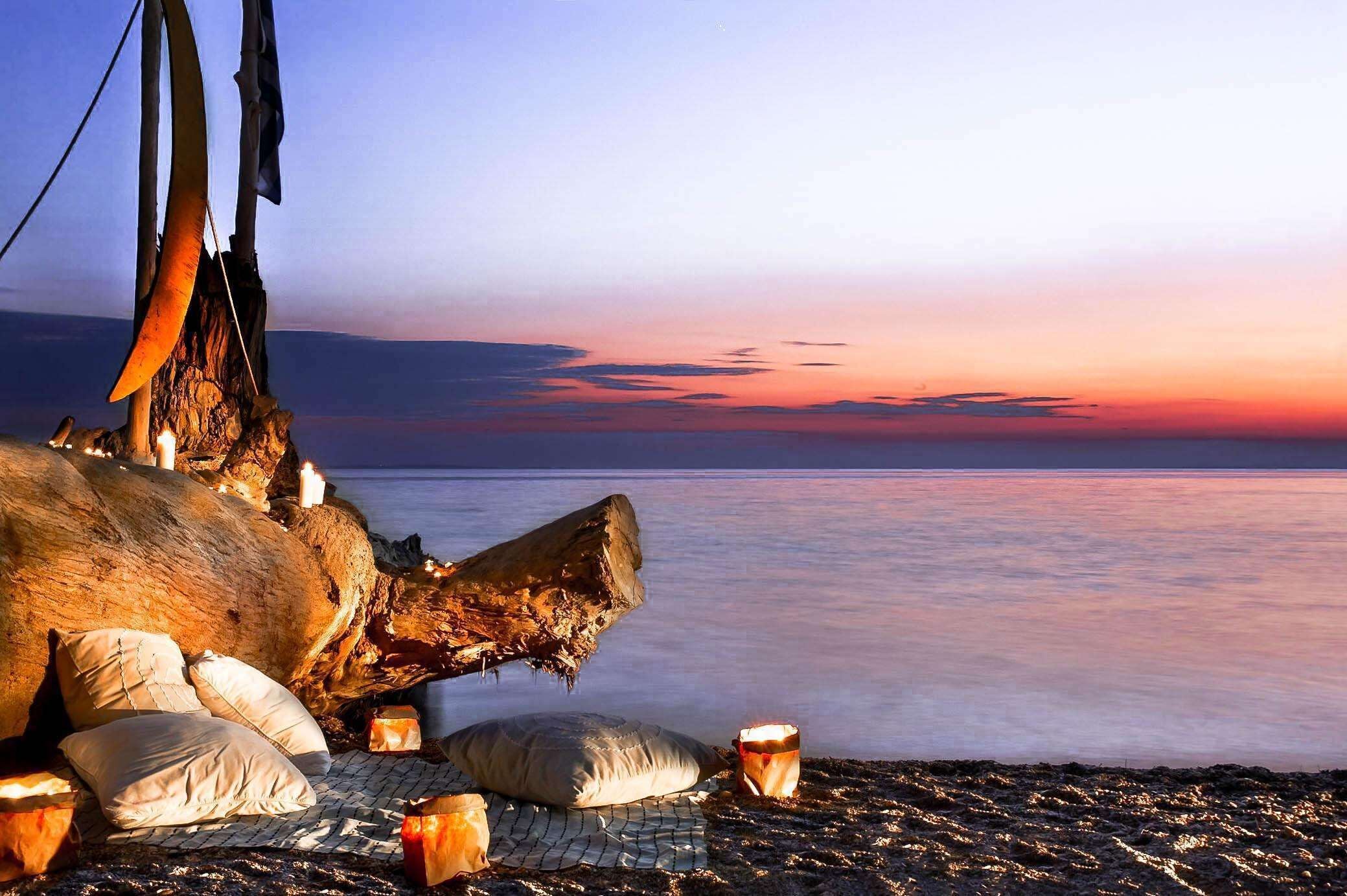 Cena de praia com um tronco grande, almofadas e velas, sugerindo um ambiente relaxante.