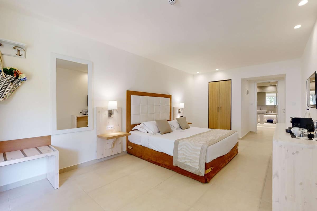 Quarto de hotel moderno com uma cama grande, mesas de cabeceira, um banco, um espelho e um banheiro visível por uma porta aberta.