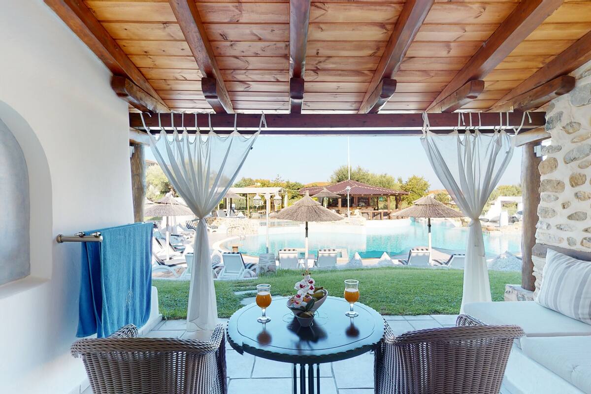 Uma área de estar coberta ao ar livre com vista para a piscina, mobiliário de vime e uma mesa com duas cervejas.