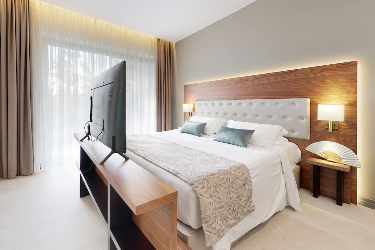 Quarto de hotel moderno com uma cama grande, televisão, cabeceira de madeira, abajures e mesa de cabeceira.