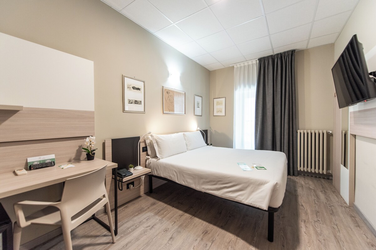Quarto de hotel com cama, escrivaninha, cadeira e televisão.