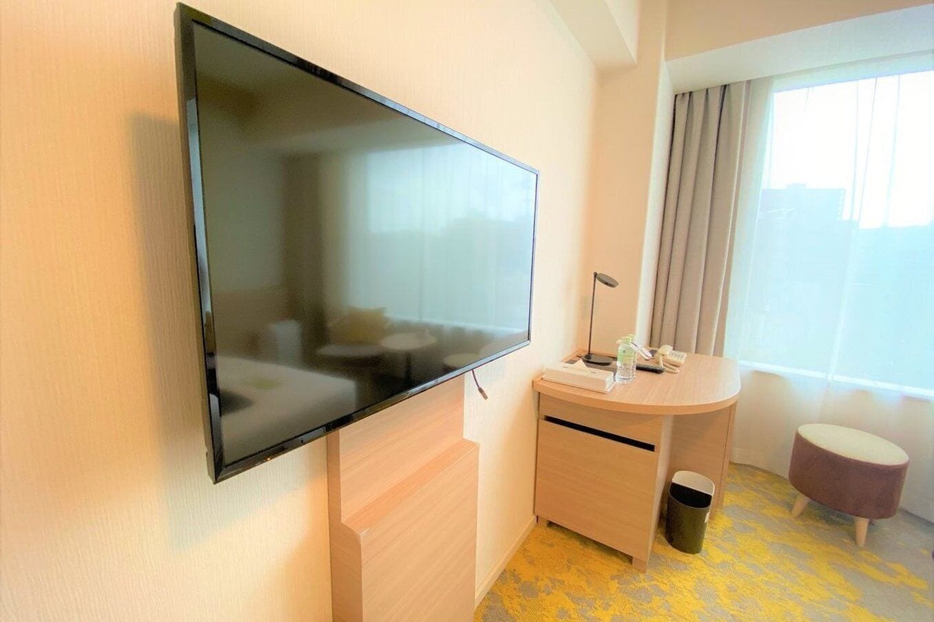Quarto de hotel com televisão de tela plana fixada na parede, uma pequena escrivaninha de madeira com luminária, um cesto de lixo e um banquinho redondo.