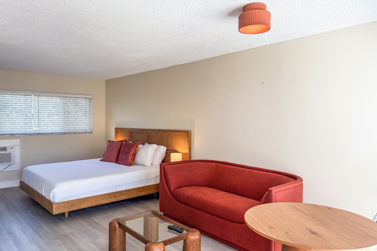 Quarto de hotel com uma cama, um sofá vermelho, uma pequena mesa de madeira e um lustre.