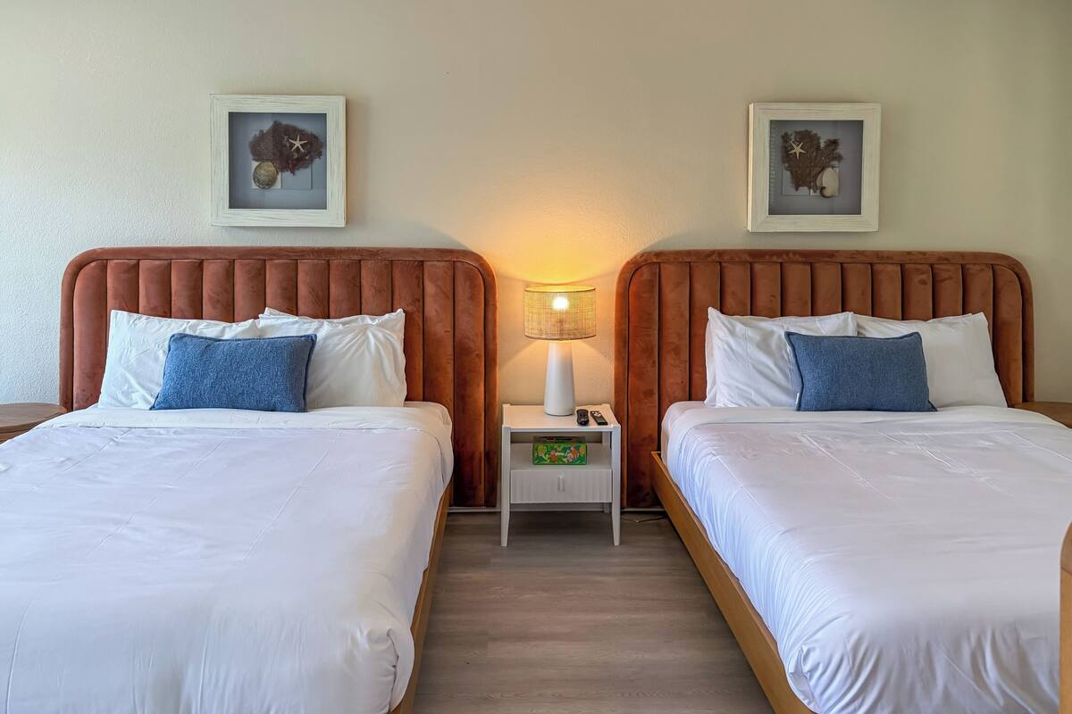 Quarto de hotel com duas camas, cada uma com roupa de cama branca e almofadas azuis, cabeceira de madeira, mesa de cabeceira com abajur e obras de arte emolduradas na parede.