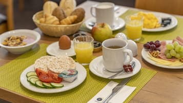 Mesa de café da manhã com croissants, pão, ovos, tomates, pepinos, iogurte, suco, café, presunto, queijo e uvas.