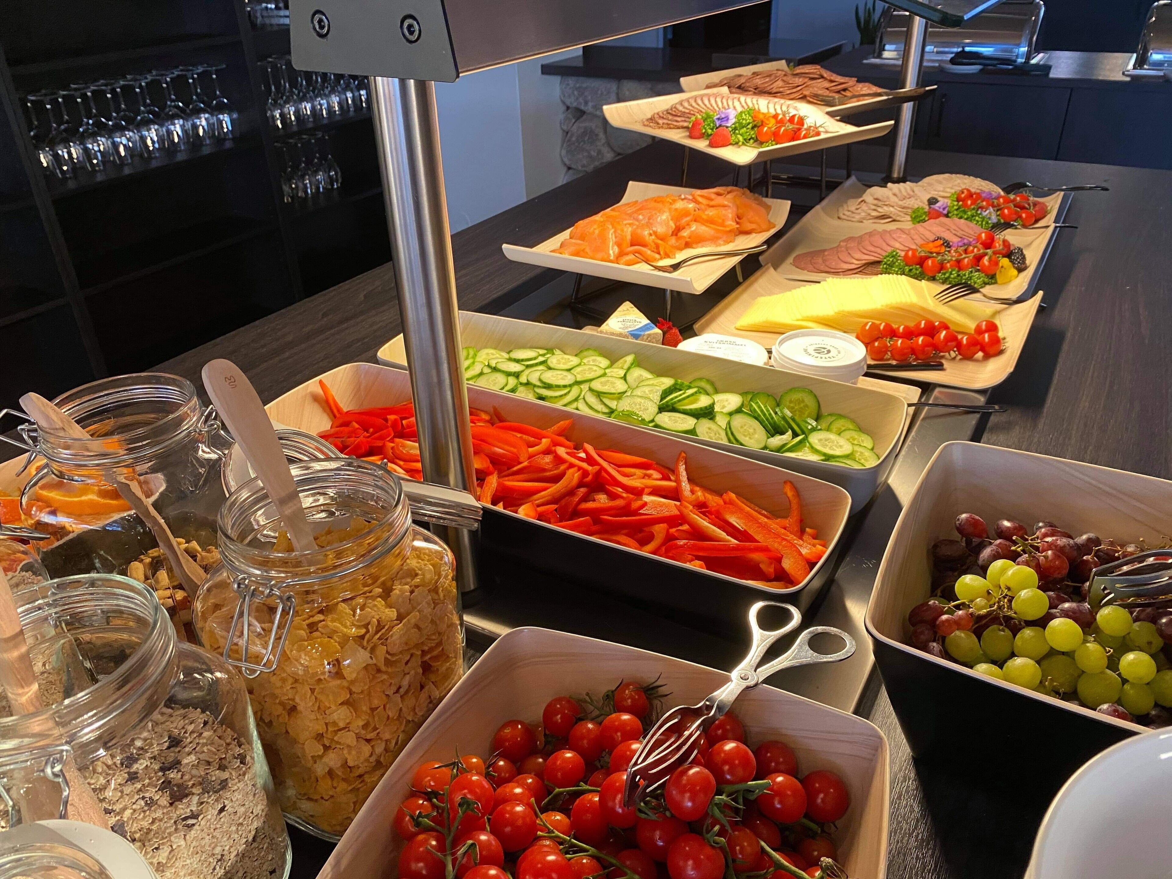 Buffet com diversas opções de comida, incluindo pepinos fatiados, tomates cereja, uvas e embutidos.
