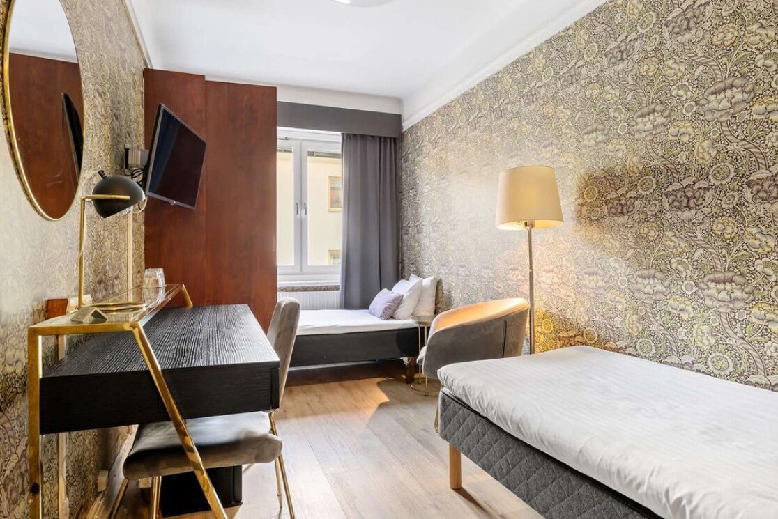 Quarto de hotel com cama, escrivaninha, cadeira, televisão na parede e janela com cortinas.
