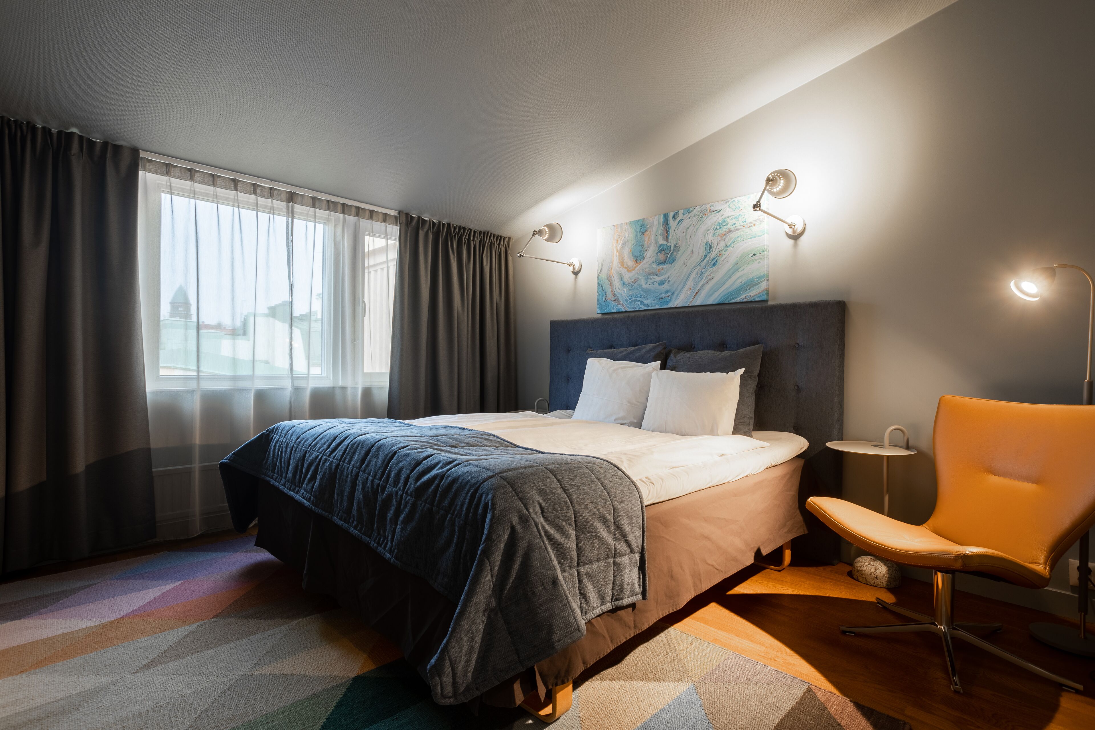 Quarto de hotel com uma cama, uma cadeira, uma mesinha de cabeceira e um quadro na parede.