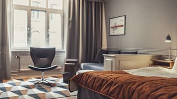 Quarto de hotel com cama, cadeira, escrivaninha, luminária e janela com cortinas.