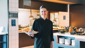 Uma pessoa com avental de chef segurando um prato de comida em uma cozinha moderna com azulejos brancos e eletrodomésticos de aço inoxidável.