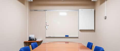 Uma sala de conferências com uma mesa comprida, cadeiras azuis, um quadro branco e uma tela de projetor.