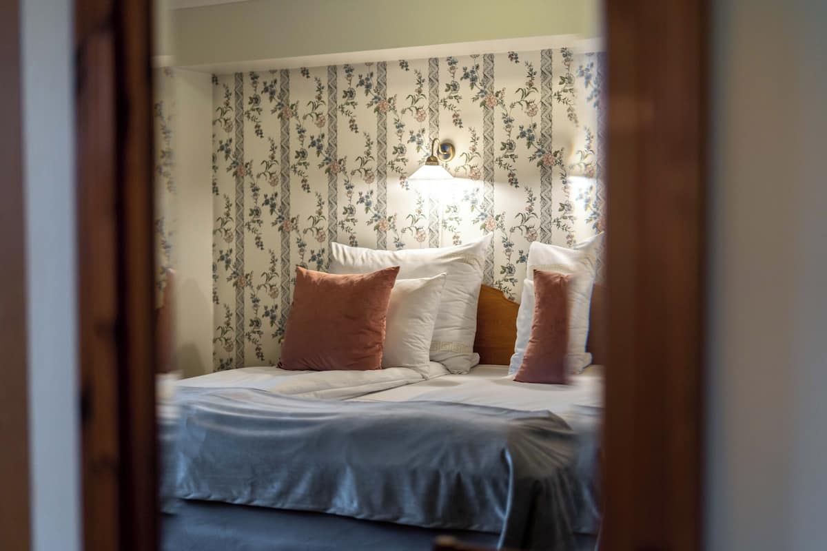 Quarto com papel de parede floral, cama com várias almofadas e mesa de cabeceira com abajur.