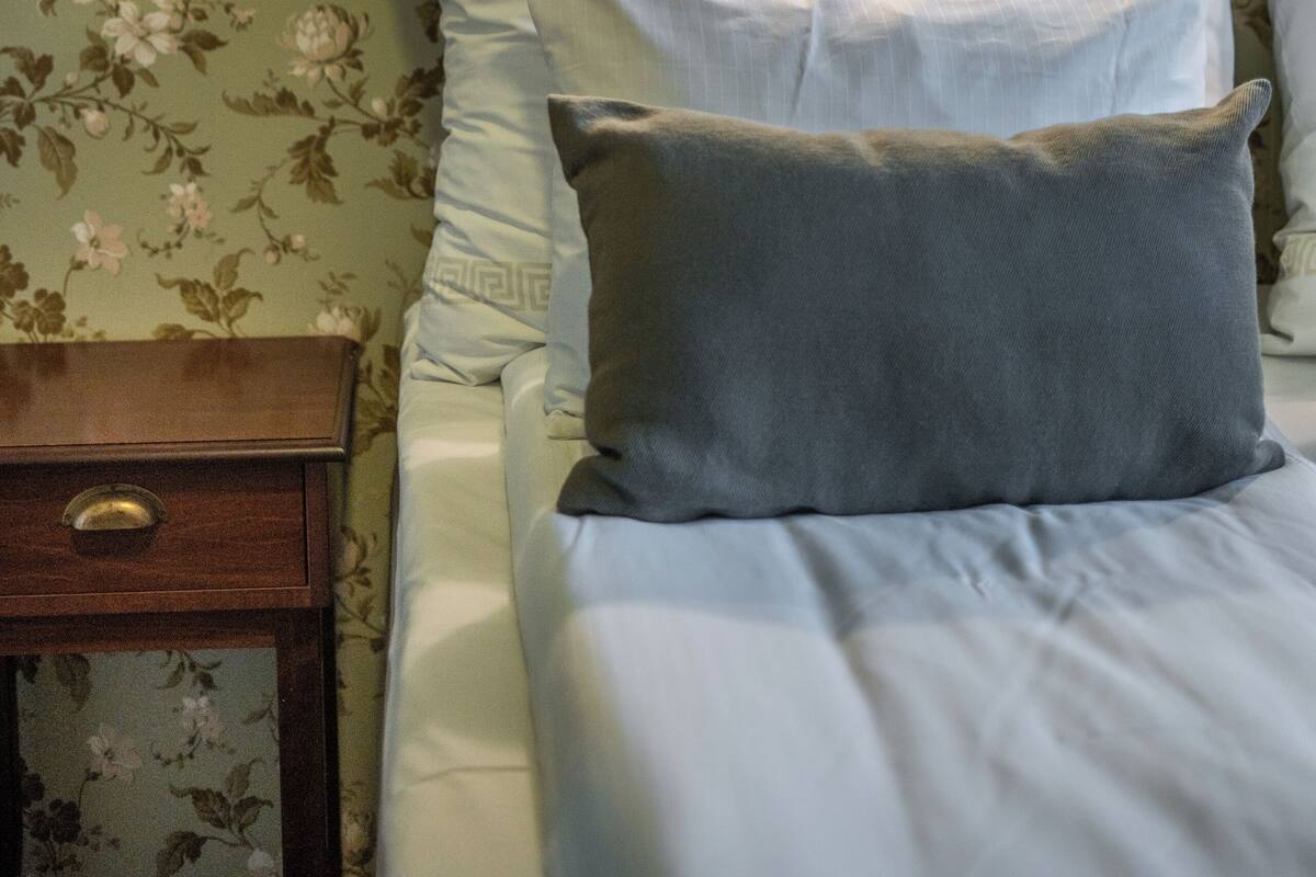 Quarto com papel de parede floral, abajur, mesa de cabeceira de madeira e cama com roupa de cama branca e cinza.