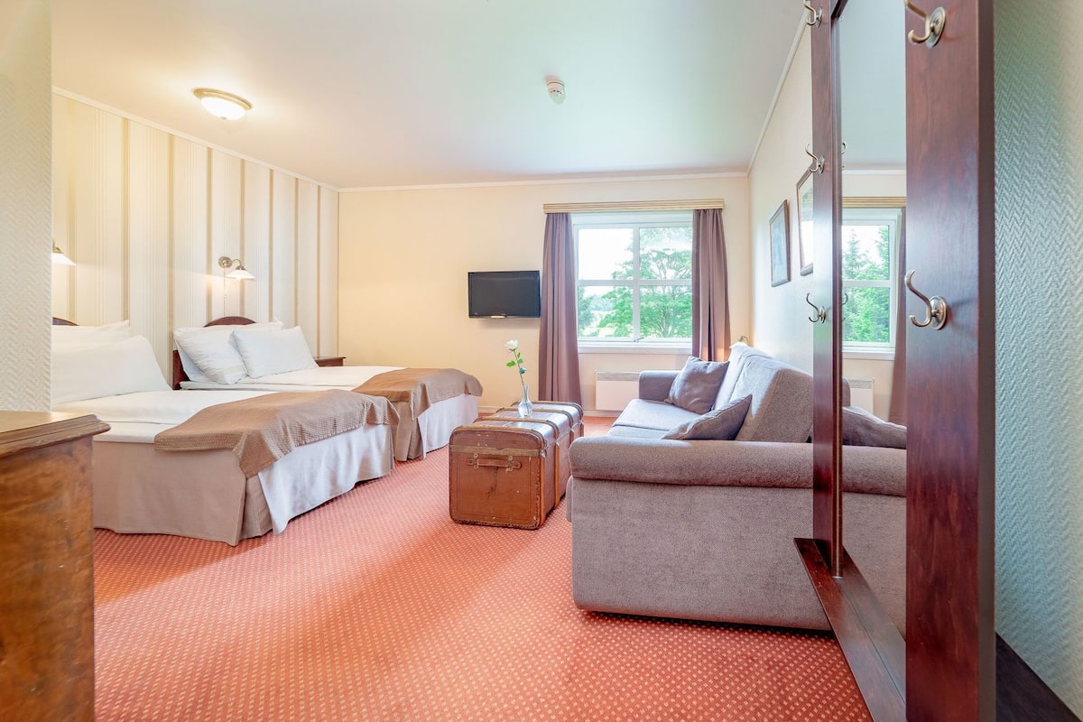 Quarto de hotel com duas camas, um sofá, uma mesa pequena, uma mala e uma televisão.