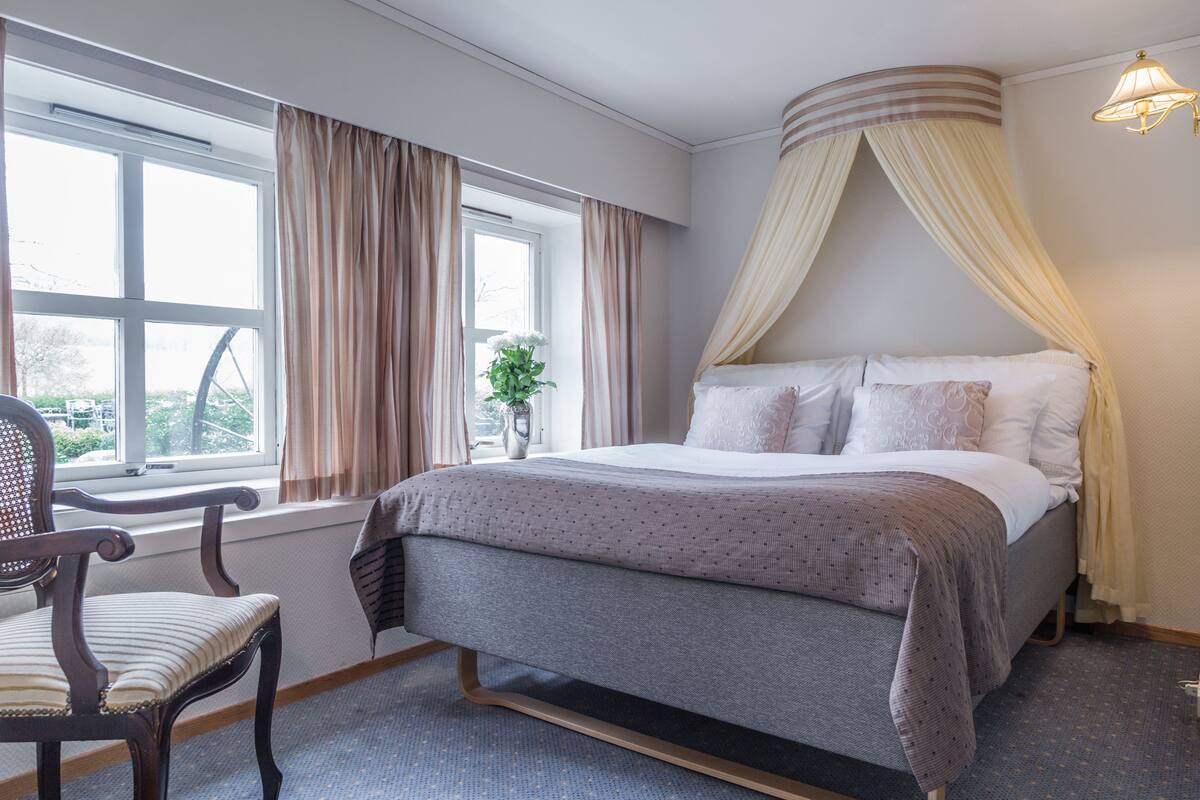 Quarto com cama, janela com cortinas, uma cadeira e abajur.