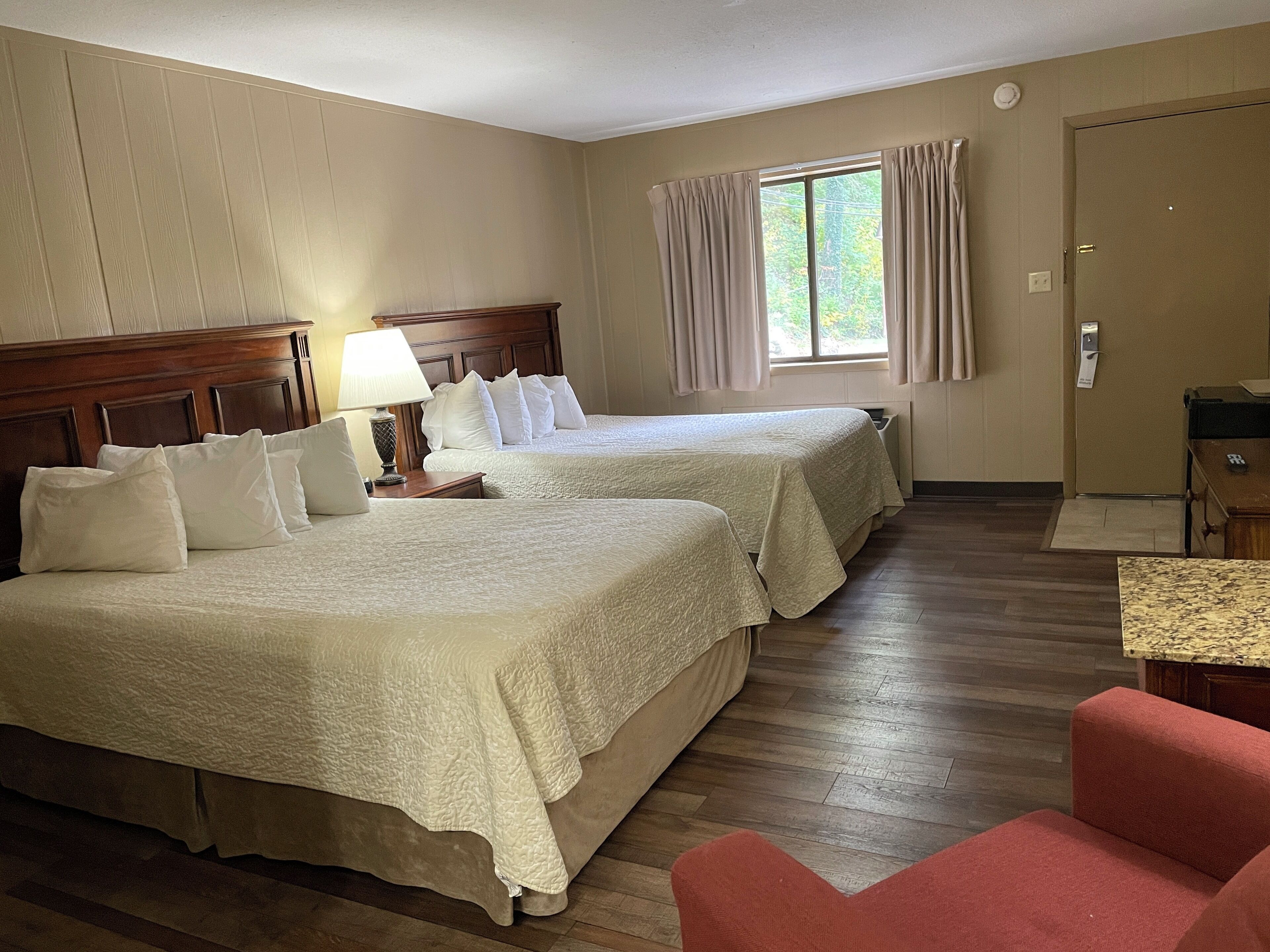 Quarto de hotel com duas camas, cabeceira de madeira, mesa de cabeceira com abajur, janela com cortinas, porta e uma pequena mesa com uma cadeira.
