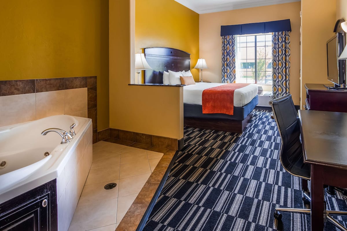 Quarto de hotel com uma banheira grande, cama com cobertor vermelho, escrivaninha com abajur e carpete estampado.