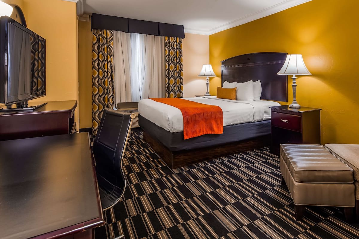 Quarto de hotel com cama, duas luminárias de cabeceira, uma escrivaninha com televisão, uma cadeira e um sofá.