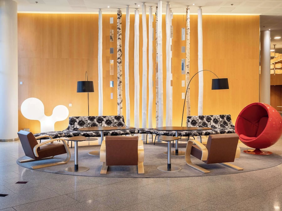 Um lobby moderno com área de estar, sofás e poltronas, incluindo uma poltrona em formato de ovo vermelha e um design de parede único com elementos verticais.