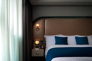 Cama bem arrumada com roupa de cama azul e branca, luminária de cabeceira e mesa de cabeceira com um telefone e um objeto pequeno.