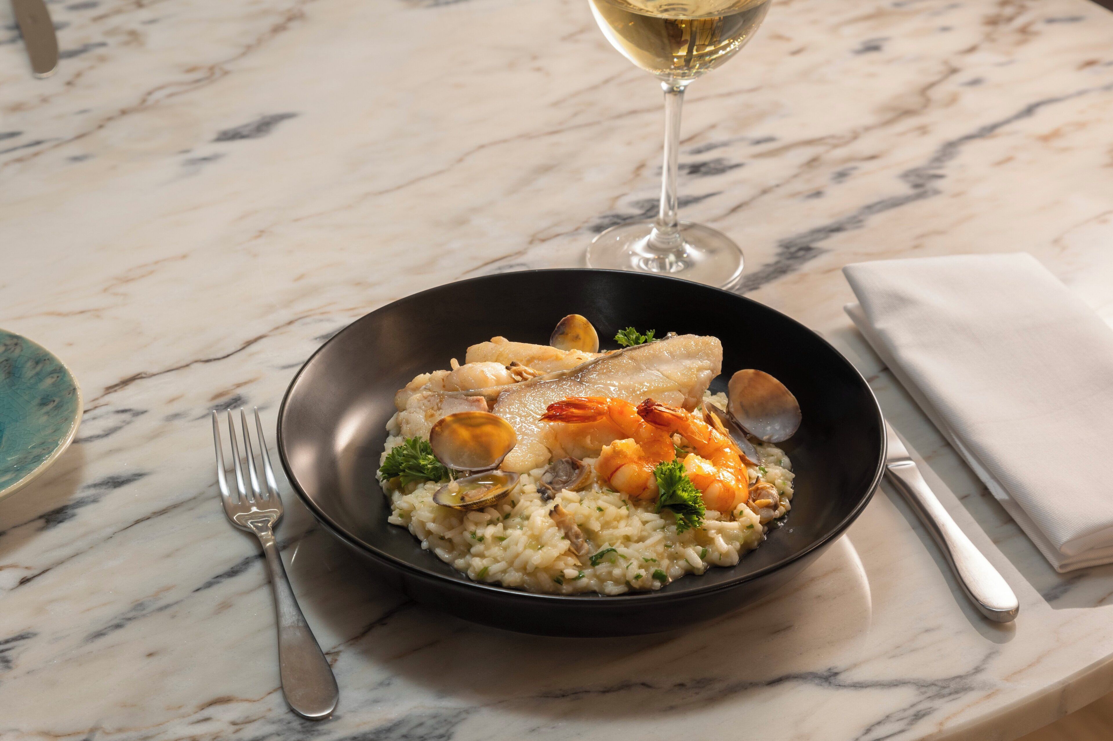 Prato de risoto de frutos do mar com amêijoas e camarões, um garfo, uma taça de vinho e um guardanapo sobre uma mesa de mármore.
