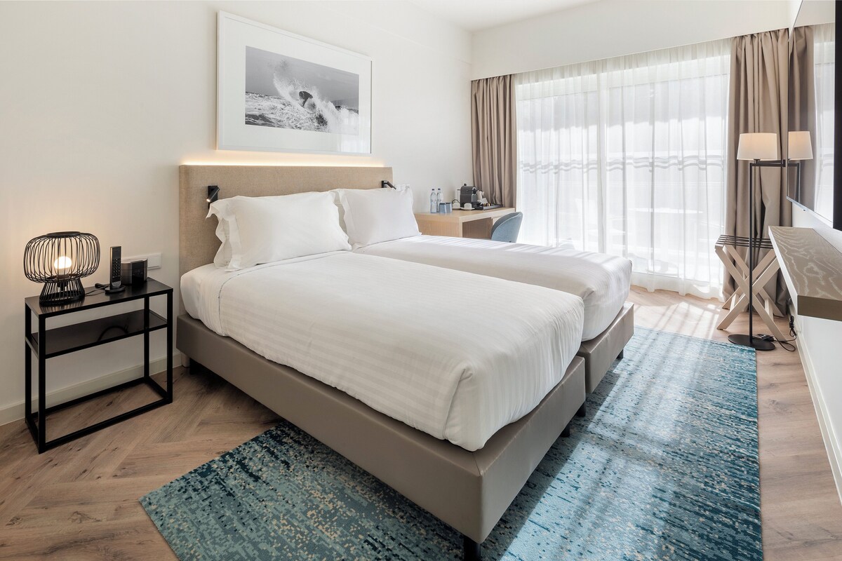 Quarto de hotel moderno com uma cama grande, mesas de cabeceira, escrivaninha e uma janela com cortinas.