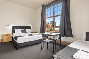 Quarto de hotel com cama, escrivaninha e cadeira. Possui janela com cortinas, mesa de cabeceira e uma pequena mesa com uma planta.