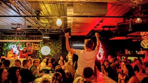 Um espaço interior lotado, com mesas e cadeiras, um palco com um artista e uma placa de neon.