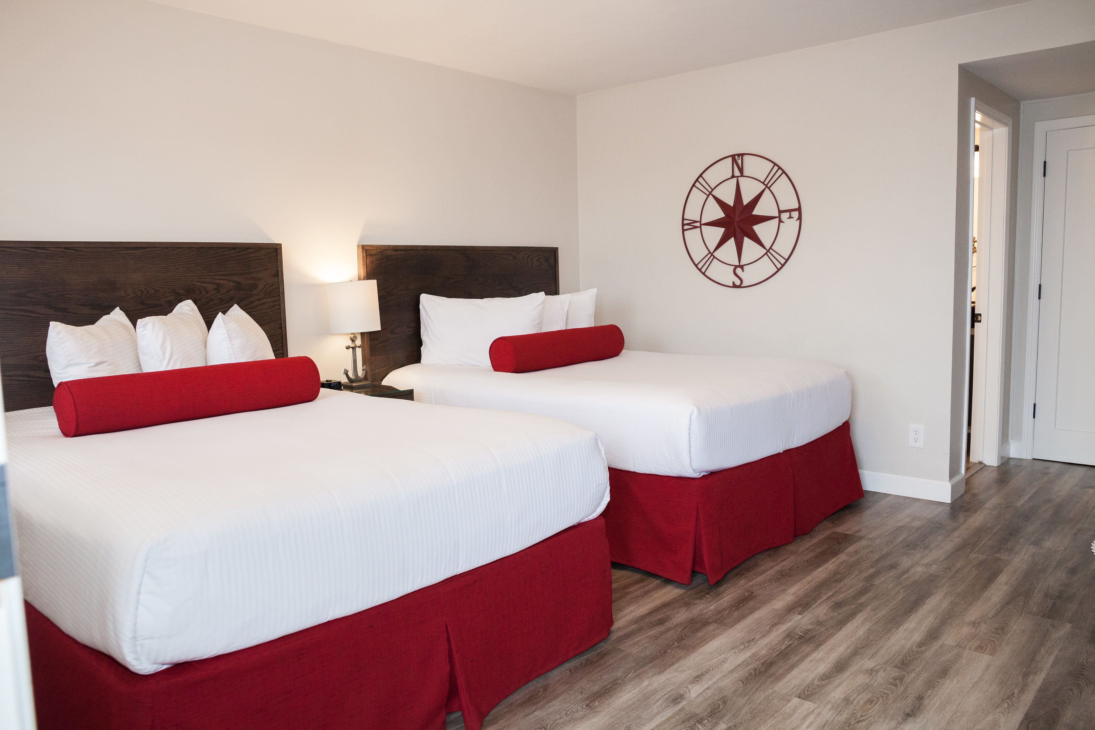 Quarto de hotel com duas camas, cada uma com roupa de cama vermelha e branca, cabeceira de madeira, relógio de parede e piso de madeira.