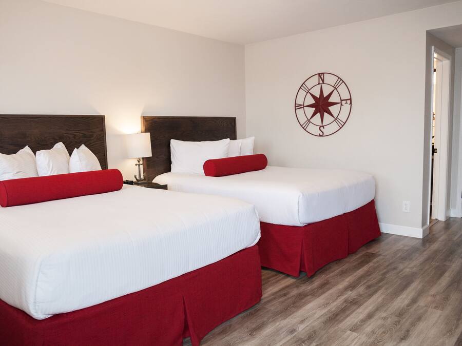 Quarto de hotel com duas camas, cada uma com roupa de cama vermelha e branca, cabeceira de madeira, relógio de parede e piso de madeira.