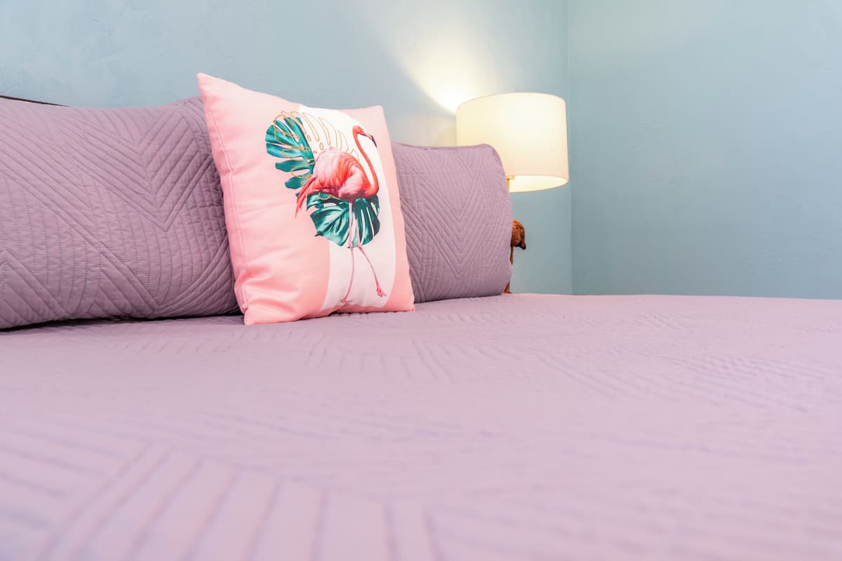 Uma cama com edredom rosa acolchoado, duas almofadas com estampa de flamingo e uma luminária fixada na parede.