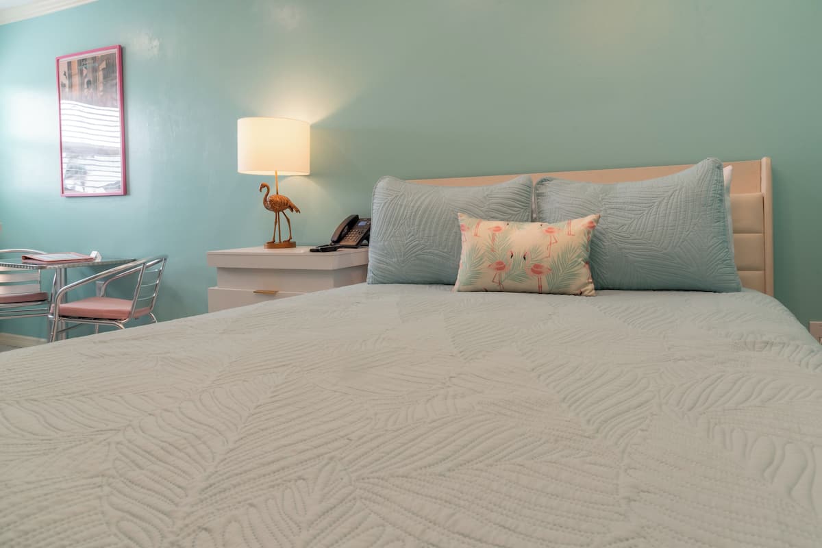 Um quarto com cama, mesa de cabeceira com luminária, cadeira e um quadro na parede.